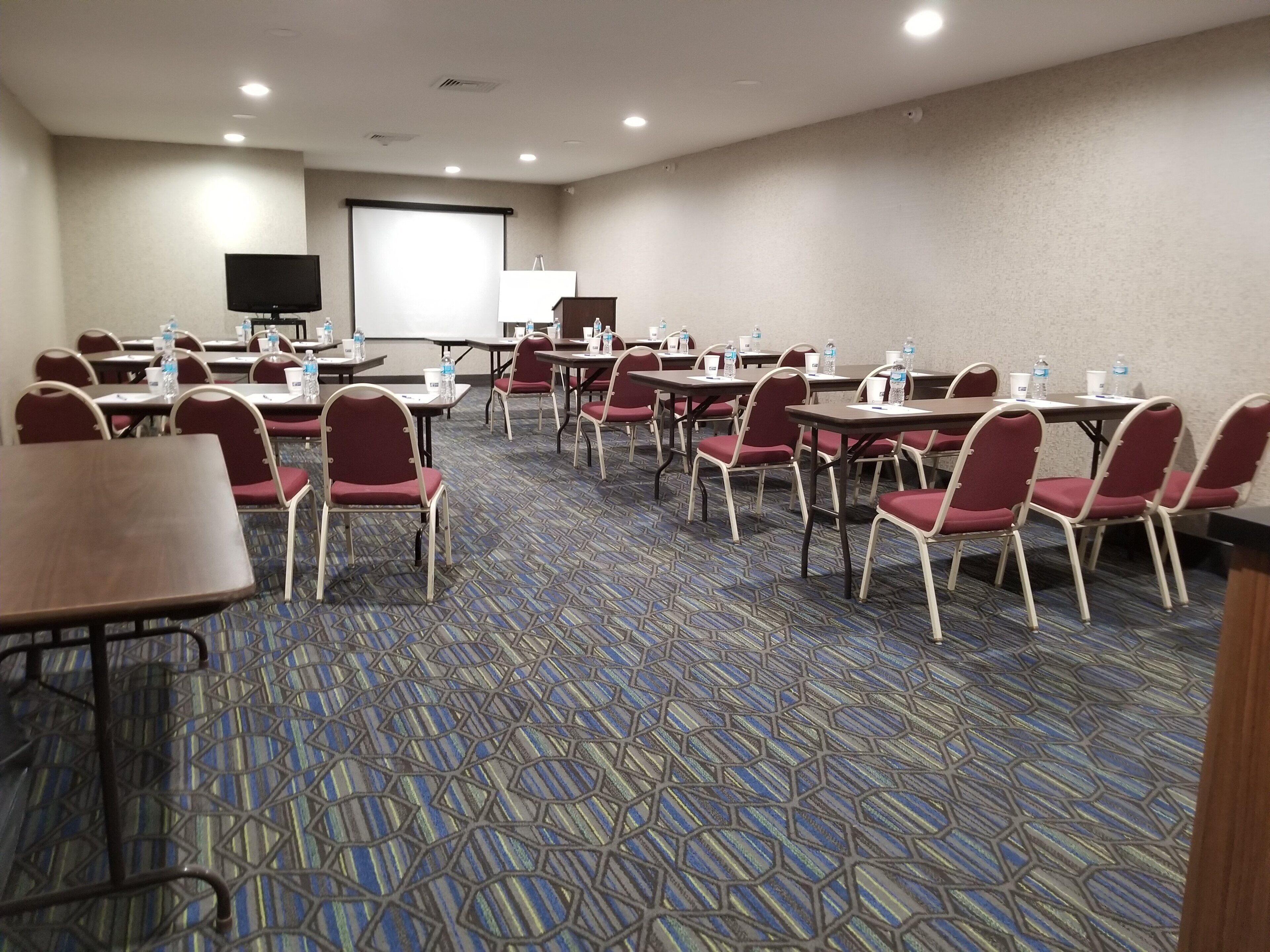 Uma sala de conferências com mesas, cadeiras, tela de projetor e piso coberto de carpete.
