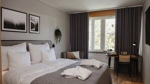 Quarto de hotel com uma cama grande, uma escrivaninha com cadeira, uma janela com cortinas e duas imagens emolduradas na parede.