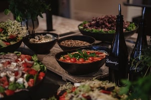 Diversos pratos sobre uma mesa de madeira, incluindo saladas, uma tigela de massa com tomates cereja e uma tigela de feijão. Também há duas garrafas de vinho na mesa.