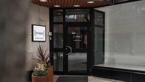 Entrada de um hotel com porta de vidro, placa indicativa e plantas em vasos.