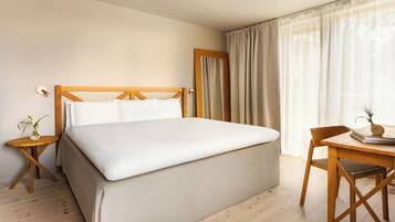 Quarto de hotel com uma cama grande, cabeceira de madeira, uma mesinha com um vaso, uma cadeira e uma janela com cortinas.