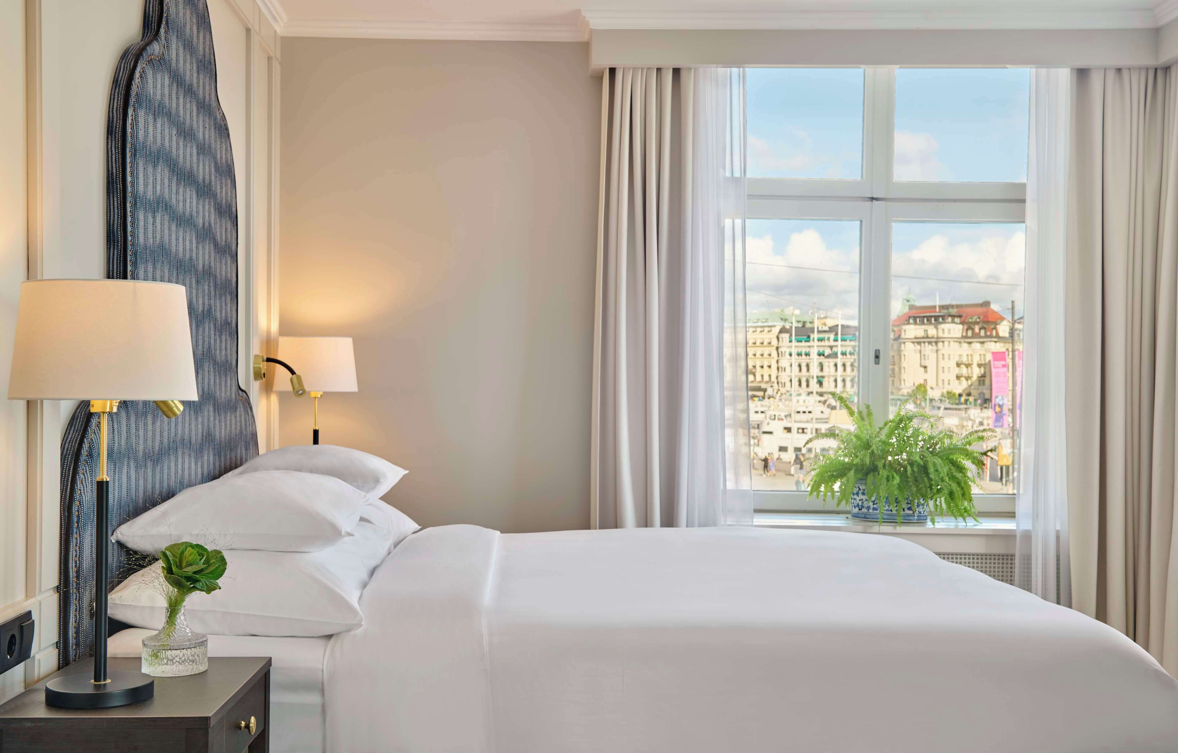 Quarto de hotel com uma cama grande, uma mesa de cabeceira com abajur, um vaso de planta e vista da cidade por uma janela ampla.