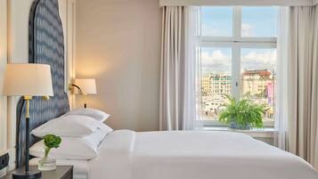Quarto de hotel com uma cama grande, uma mesa de cabeceira com abajur, um vaso de planta e vista da cidade por uma janela ampla.