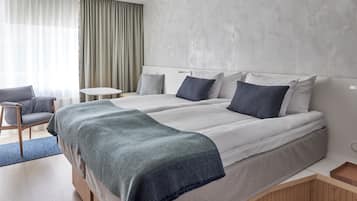 Quarto de hotel moderno com duas camas, uma mesinha, uma cadeira e vista para o exterior.