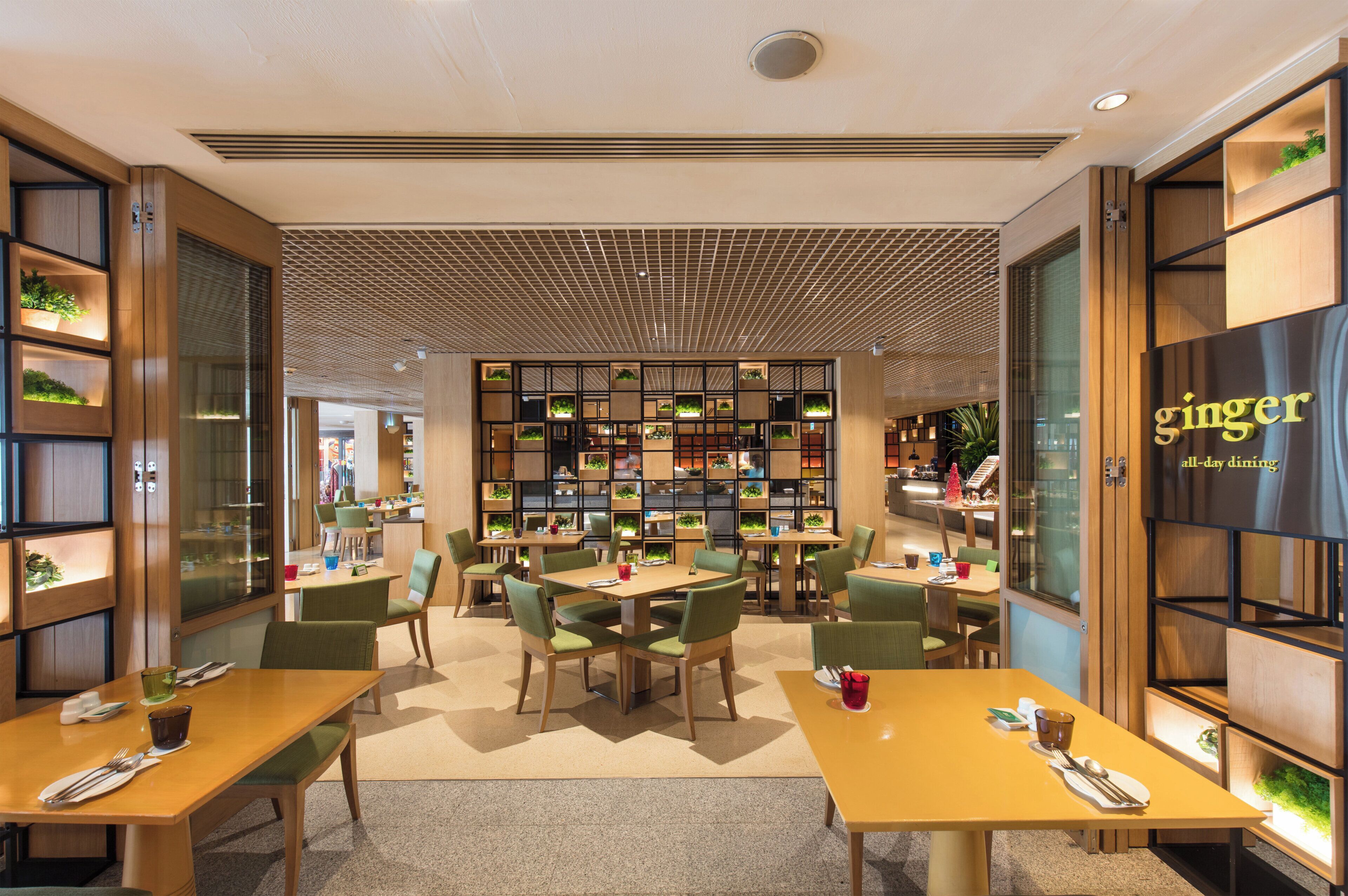 Interior de restaurante com mesas e cadeiras de madeira, teto com padrão de grade e prateleiras com plantas.
