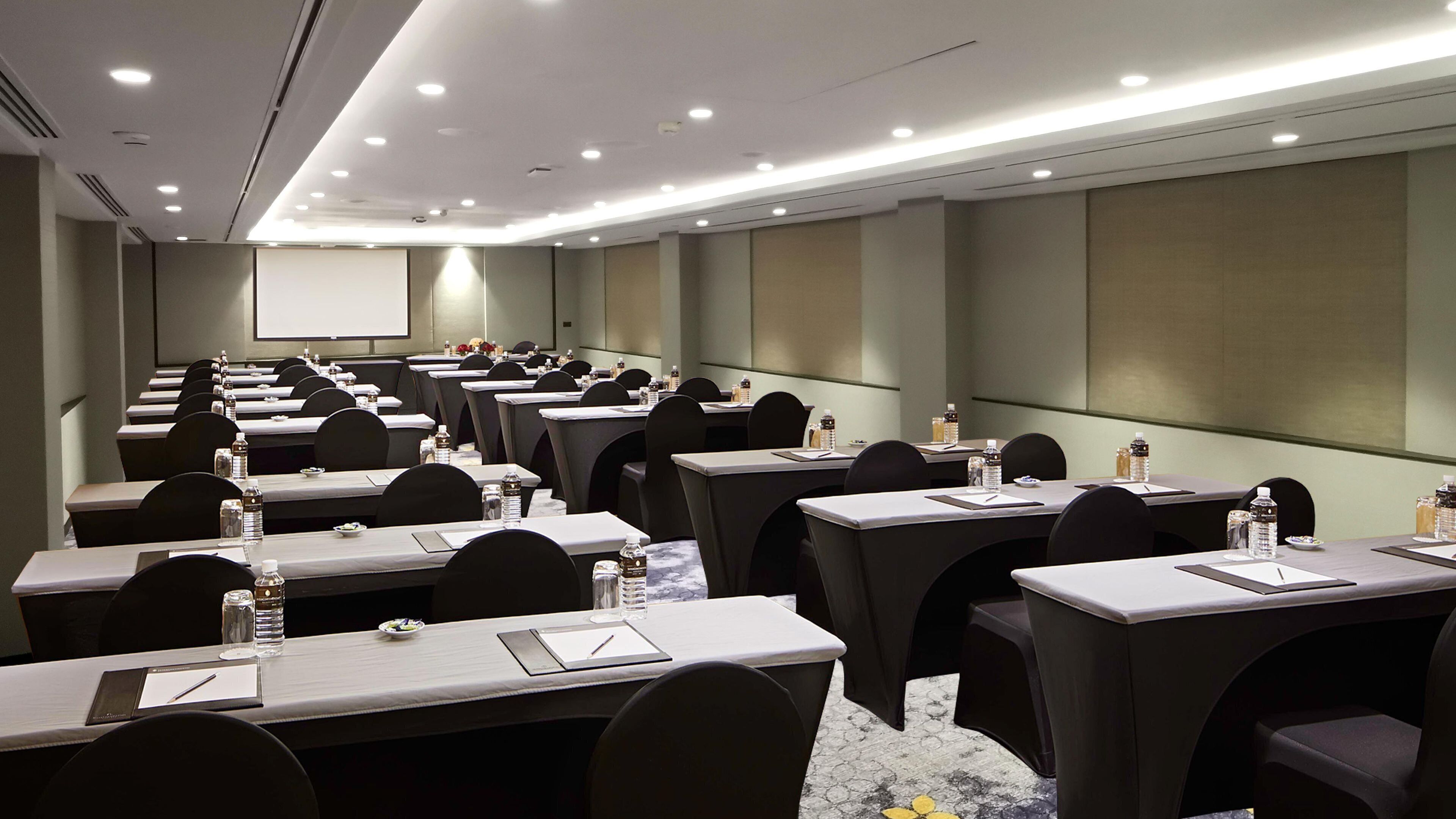 Uma sala de conferências com fileiras de mesas, cadeiras e uma tela de projeç ão.