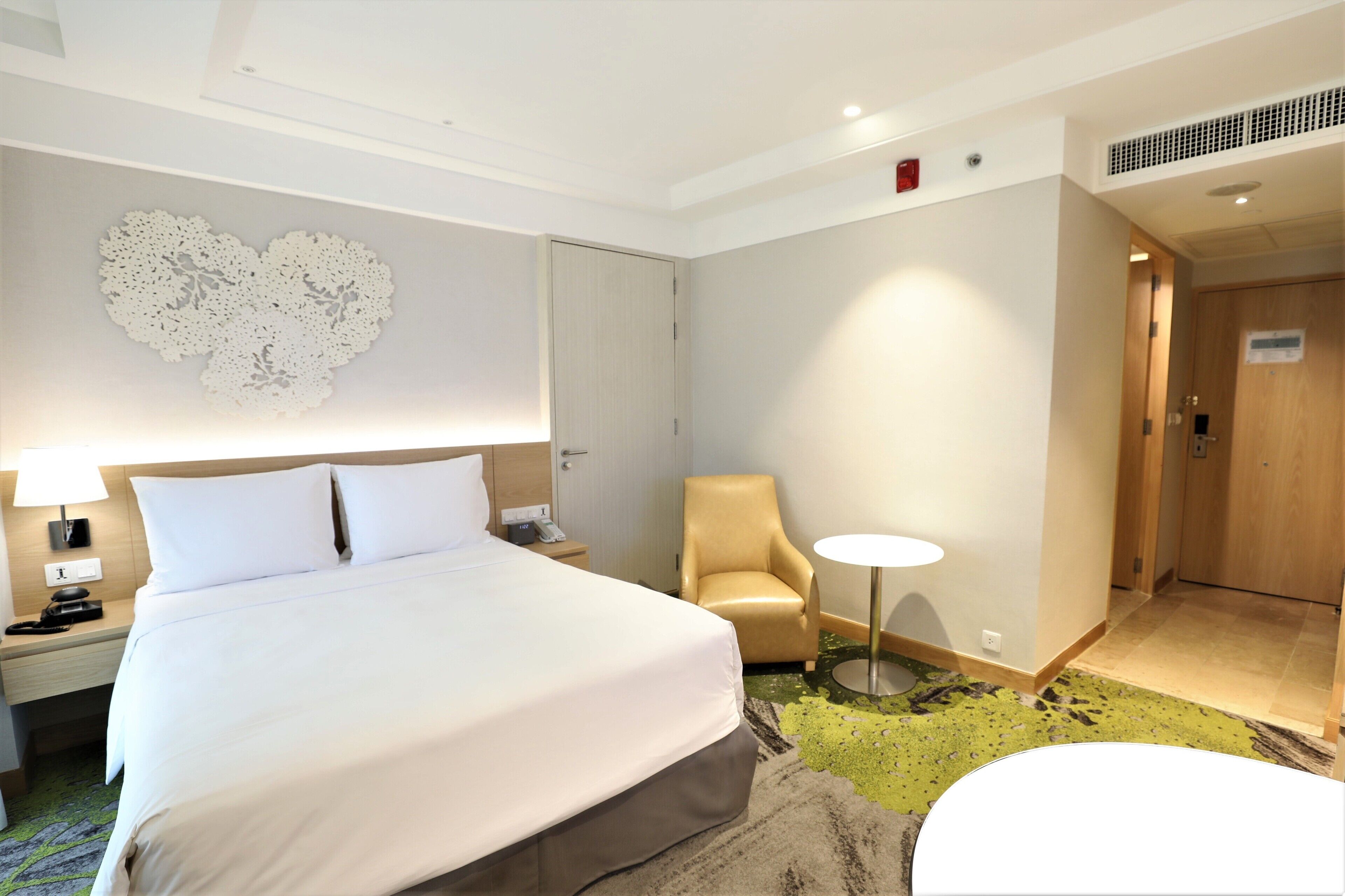 Quarto de hotel com cama grande, uma cadeira, uma mesa pequena e um objeto decorativo na parede.
