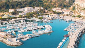 Uma marina com vários barcos atracados, cercada por edifícios residenciais e um trecho costeiro rochoso.