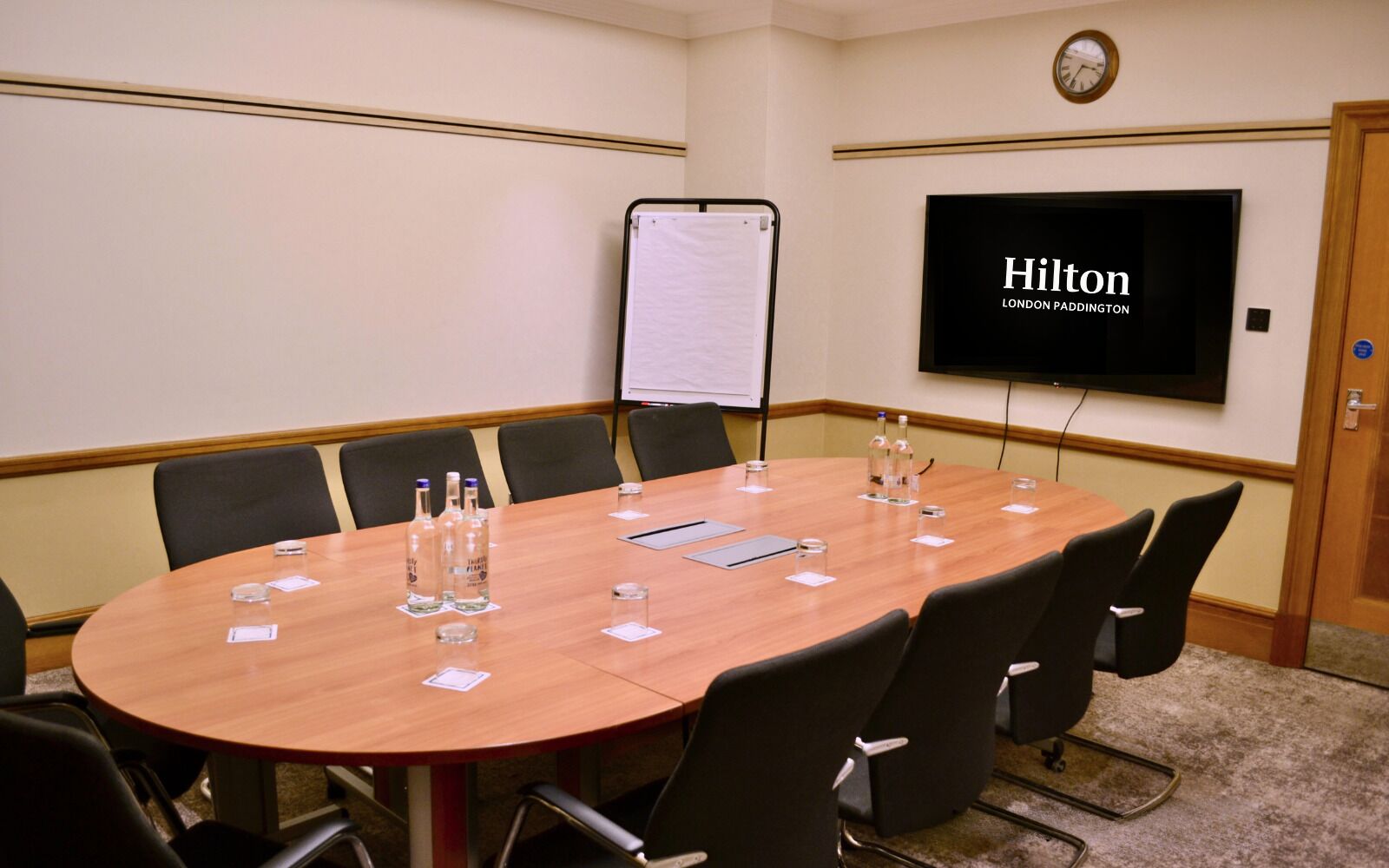 Uma sala de conferências com uma grande mesa oval, cadeiras pretas, garrafas de água, placas de identificação, um quadro branco e uma tela de televisão exibindo "Hilton London Paddington".