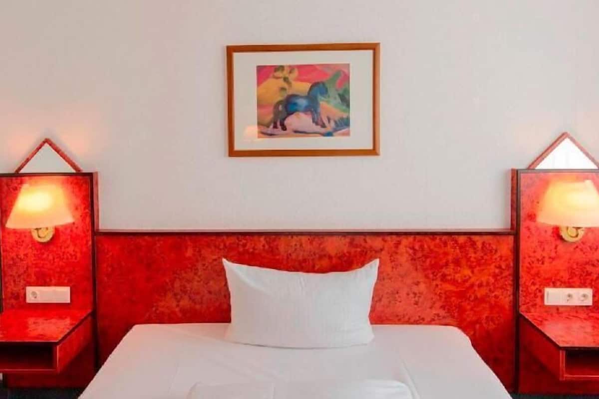 Uma cama com roupa de cama branca, duas mesas de cabeceira com luminárias, um painel de cabeceira vermelho e um quadro pendurado na parede.