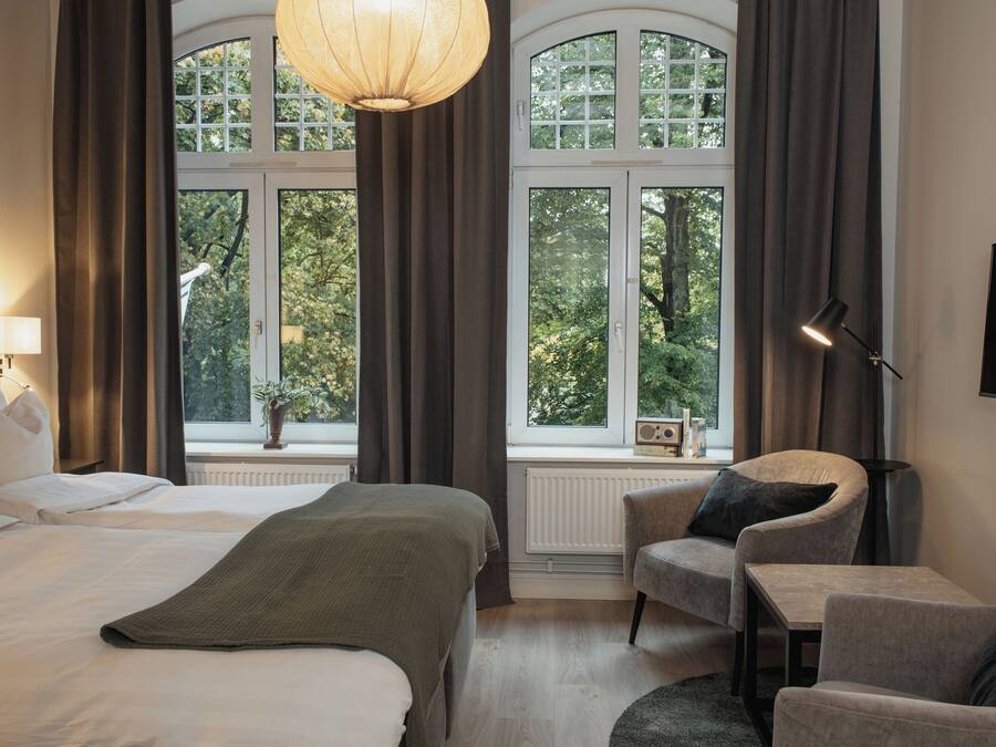 Quarto de hotel com uma cama grande, duas poltronas, uma mesinha, um abajur e vista para árvores através de amplas janelas.