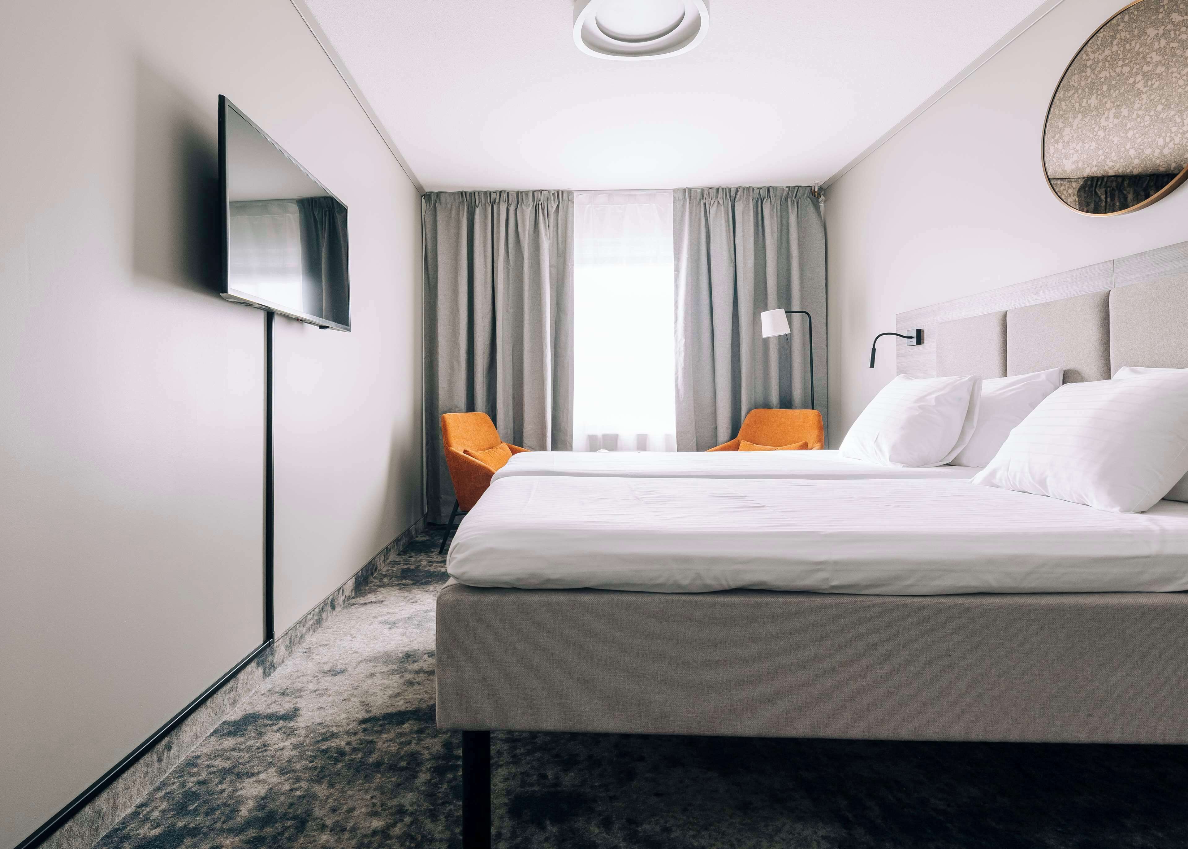 Quarto de hotel com cama, duas cadeiras, televisão, luminária e janela com cortinas.