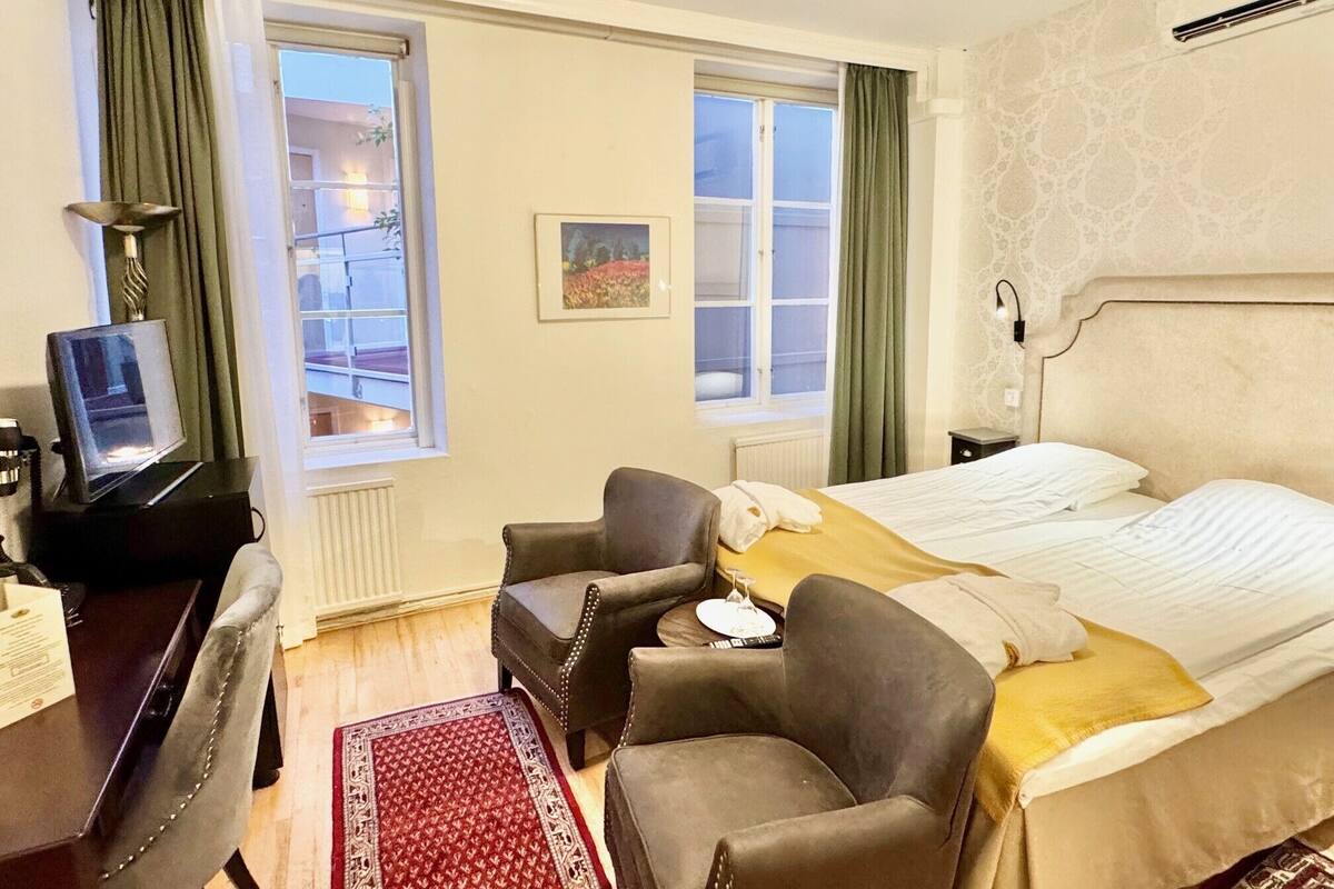 Quarto de hotel com cama, duas poltronas, uma escrivaninha com televisão, janela com cortinas e radiador.
