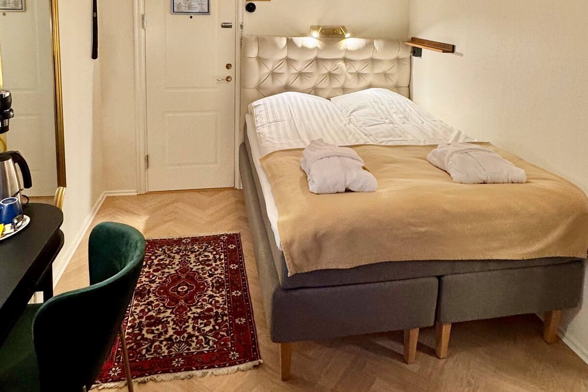 Quarto compacto com cama, mesa de cabeceira, cadeira e luminária pendente.