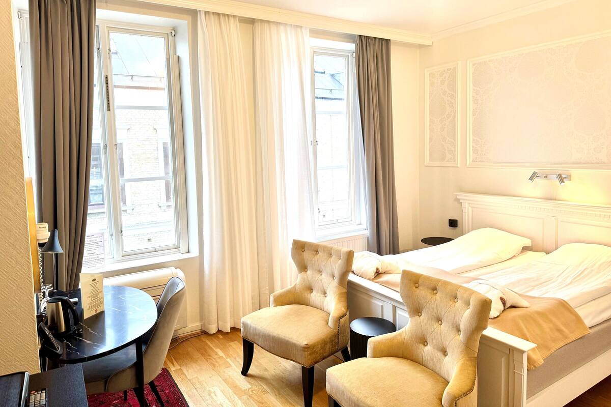 Quarto de hotel com uma cama, duas poltronas, uma mesinha e uma janela com cortinas.