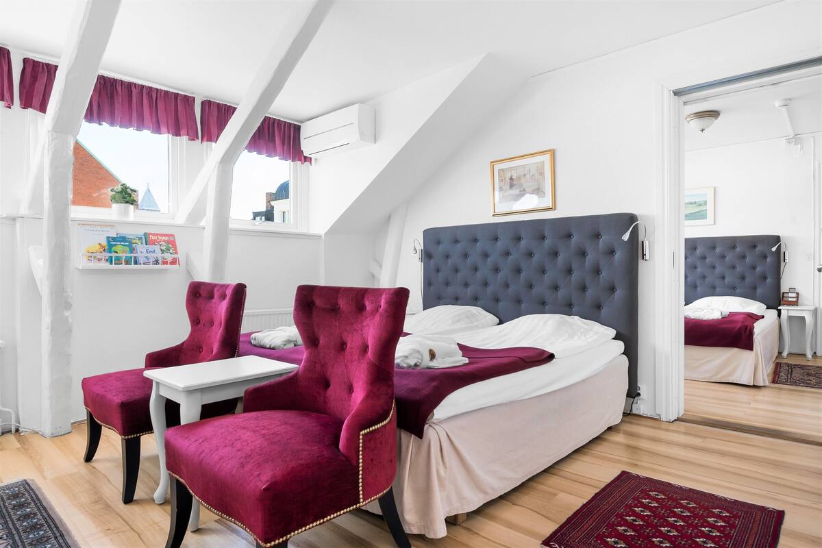 Quarto com uma cama grande, duas poltronas de veludo vermelho, uma mesinha branca e uma janela com vista para um edifício.