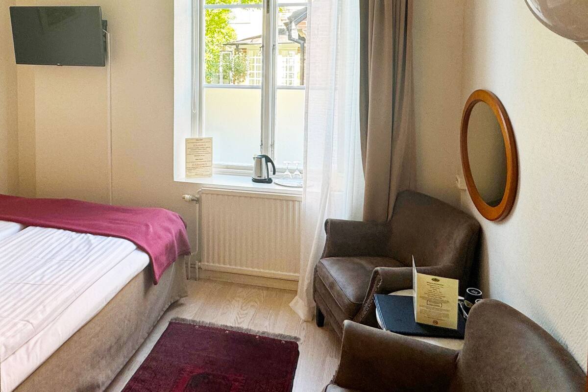 Quarto de hotel com cama, cadeira, janela com cortinas, radiador e televisão presa à parede.