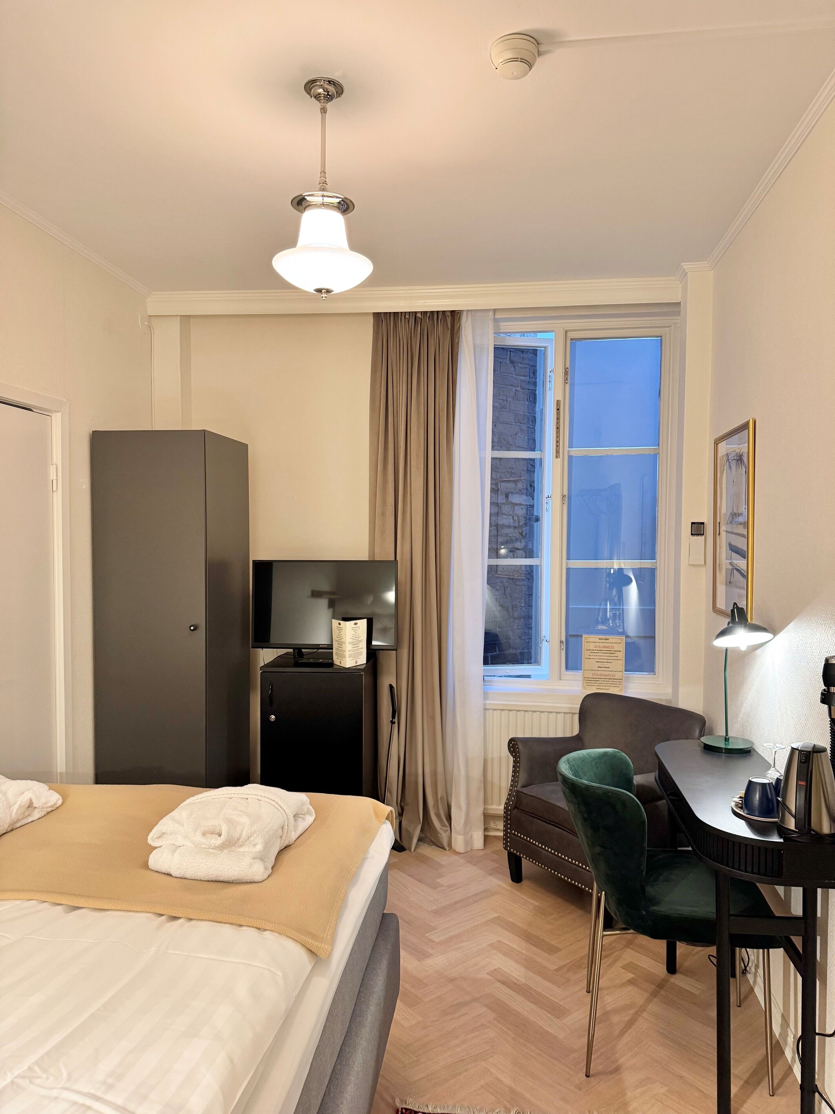 Quarto de hotel com cama, escrivaninha, cadeira, guarda-roupa e janela com cortinas.