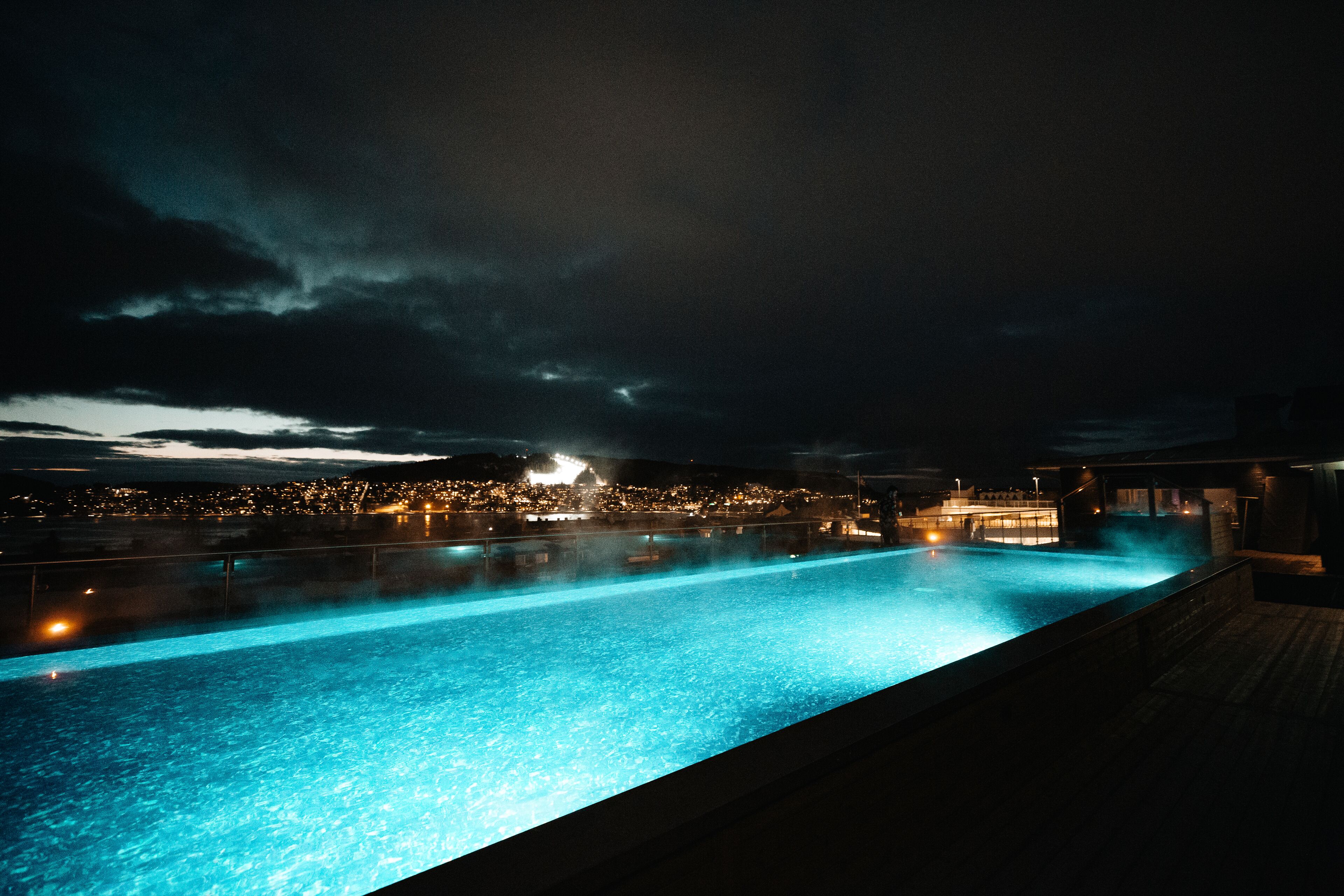 Uma piscina à noite com luzes da cidade ao fundo.