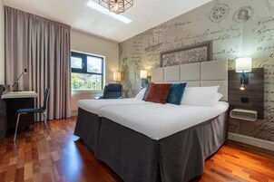 Quarto de hotel com uma cama grande, uma escrivaninha e uma cadeira. Há uma janela com cortinas, uma parede com detalhes decorativos e uma luminária na mesa lateral.