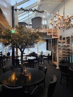 Um espaço interior moderno com uma árvore central decorada com luzes, rodeada por mesas e cadeiras.