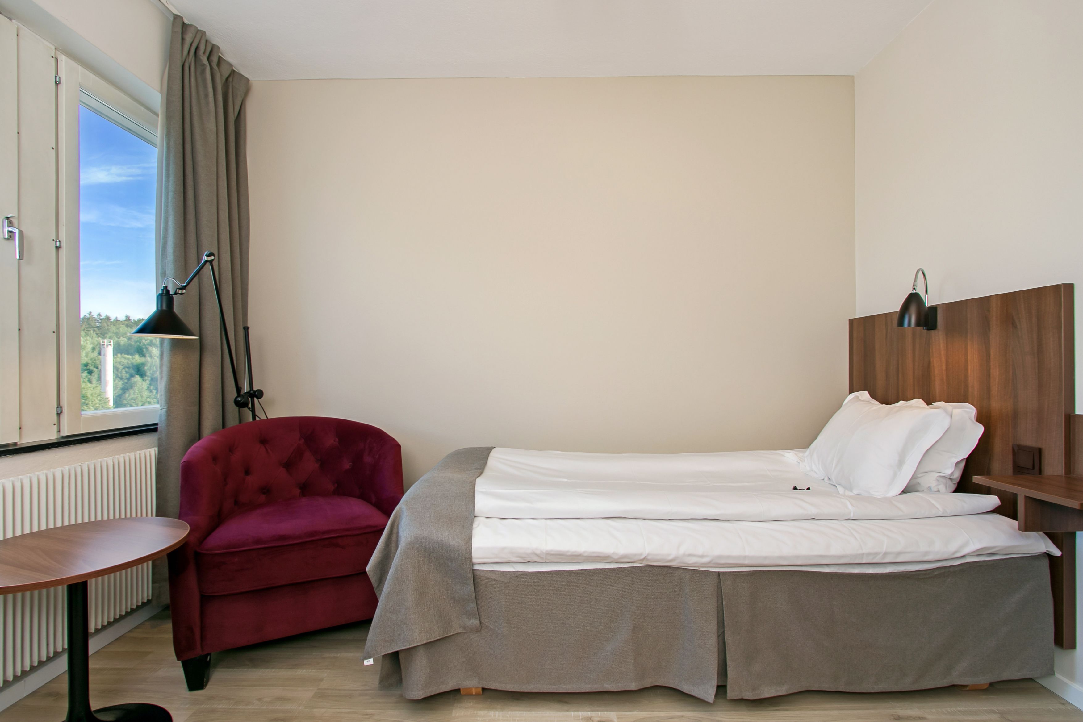Quarto com uma cama de solteiro, uma poltrona vermelha, uma mesa e uma janela com cortinas.