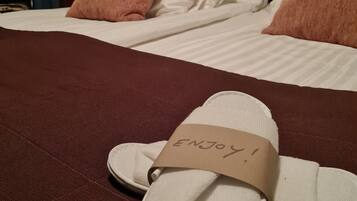 Uma cama com roupa de cama branca e castanha, uma etiqueta castanha num par de chinelos brancos e uma parede com padrão floral.