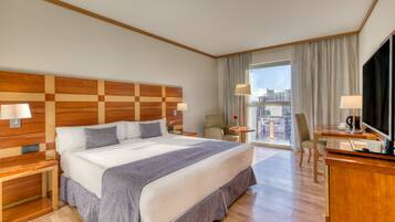 Quarto de hotel com cama grande, cabeceira de madeira, escrivaninha com TV e vista da cidade por grandes janelas.