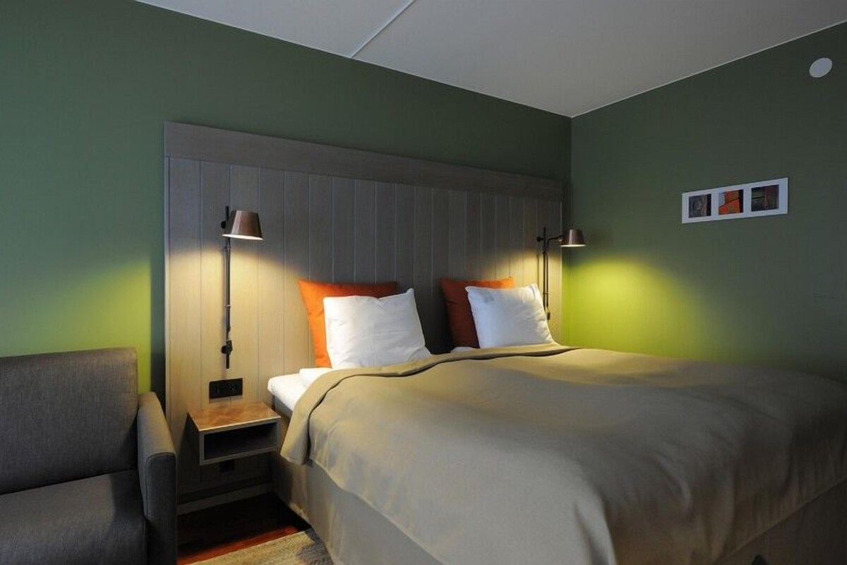 Quarto de hotel com uma cama, mesas de cabeceira, uma cadeira e uma prateleira na parede.