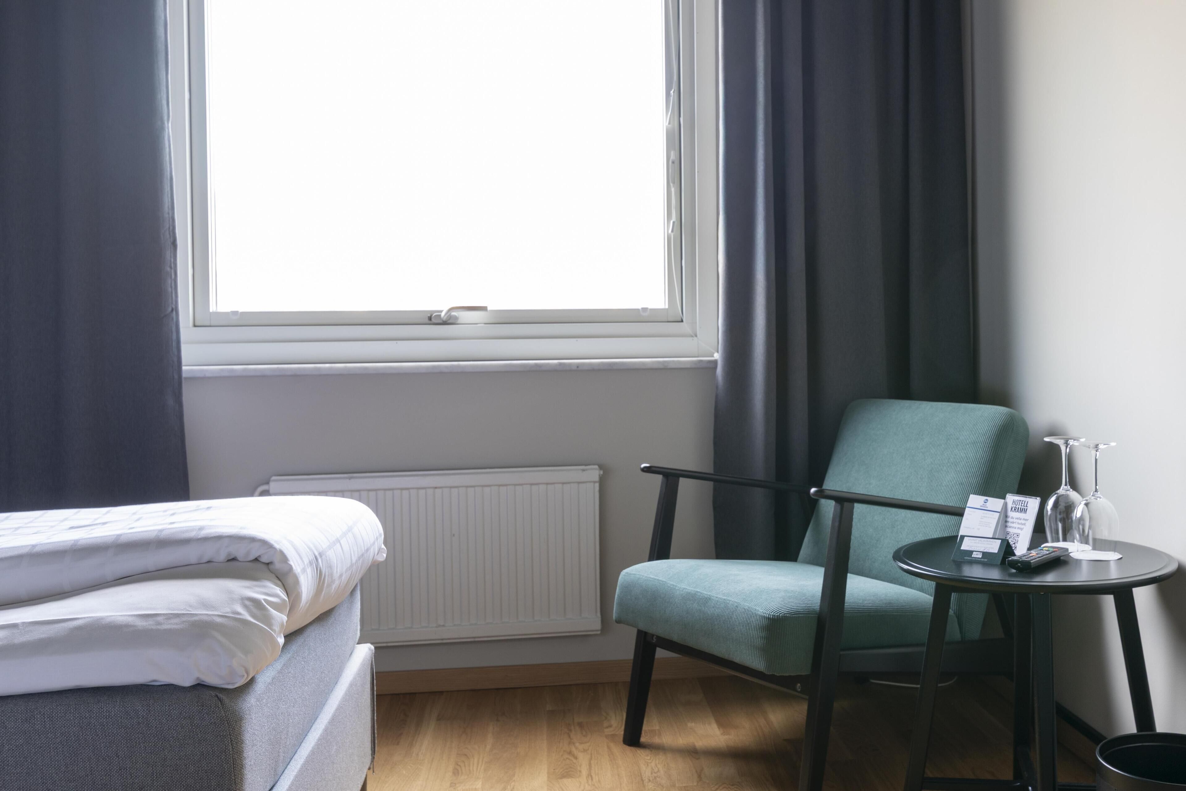 Quarto de hotel com uma cama, uma cadeira, uma mesinha com um copo, um controle remoto e uma placa.