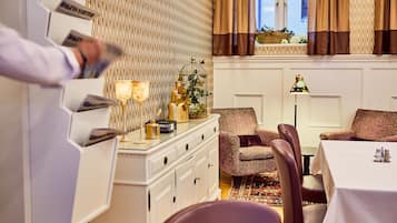 Quarto de hotel com uma receção, área de refeições com mesa e cadeiras, e uma planta decorativa no parapeito da janela.