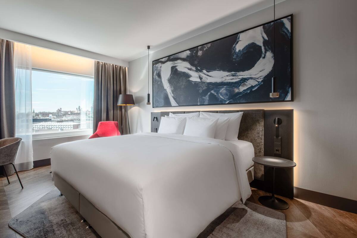 Quarto de hotel moderno com uma cama grande, vista para a cidade, uma cadeira vermelha e um quadro abstrato de grandes dimensões na parede.