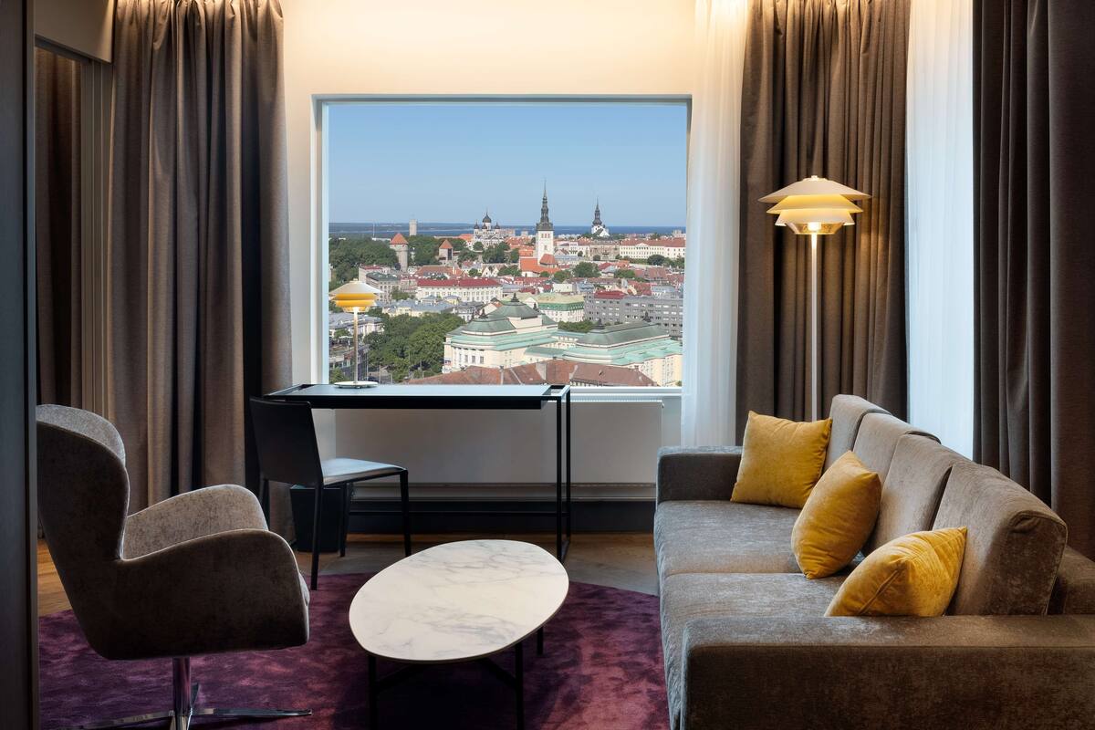 Quarto de hotel moderno com uma janela grande que oferece vista da cidade, um sofá com almofadas amarelas, uma mesa pequena e uma cadeira.