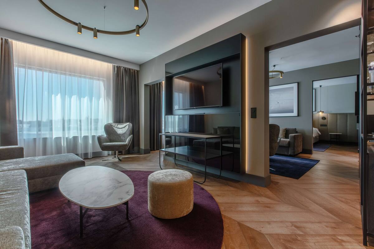 Uma sala de estar moderna com sofá, pufê e mesa de centro, uma televisão de tela plana na parede e uma área de jantar com mesa e cadeiras de vidro.