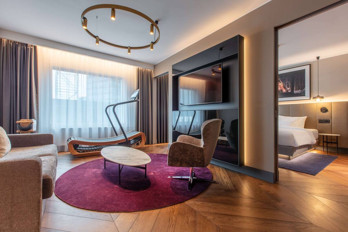 Quarto de hotel moderno com uma televisão grande, um sofá, uma poltrona, uma mesinha e uma lareira.