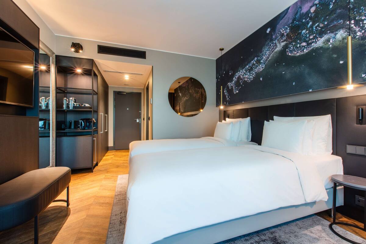 Quarto de hotel moderno com uma cama grande, área de estar, cozinha planejada e um mural com vista para a cidade na parede.