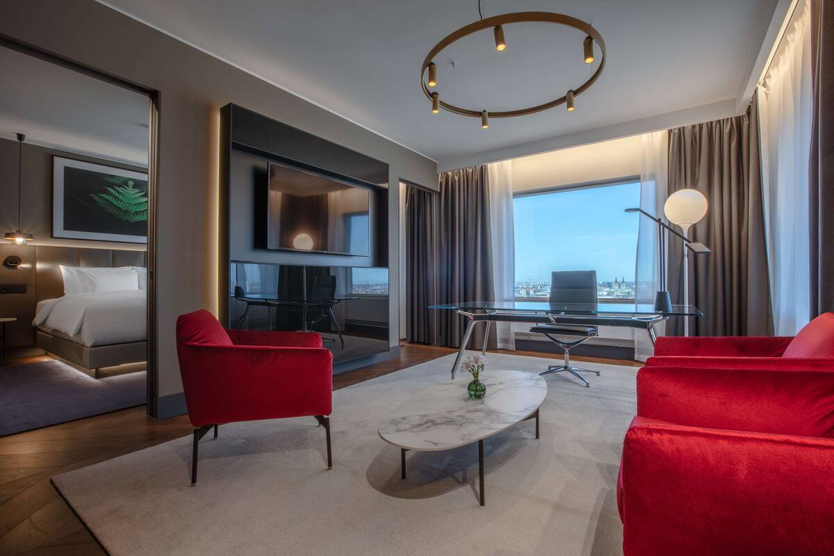 Quarto de hotel moderno com cama, poltronas vermelhas, mesa de centro de mármore, escrivaninha com cadeira, uma janela grande com cortinas e luminária no teto.