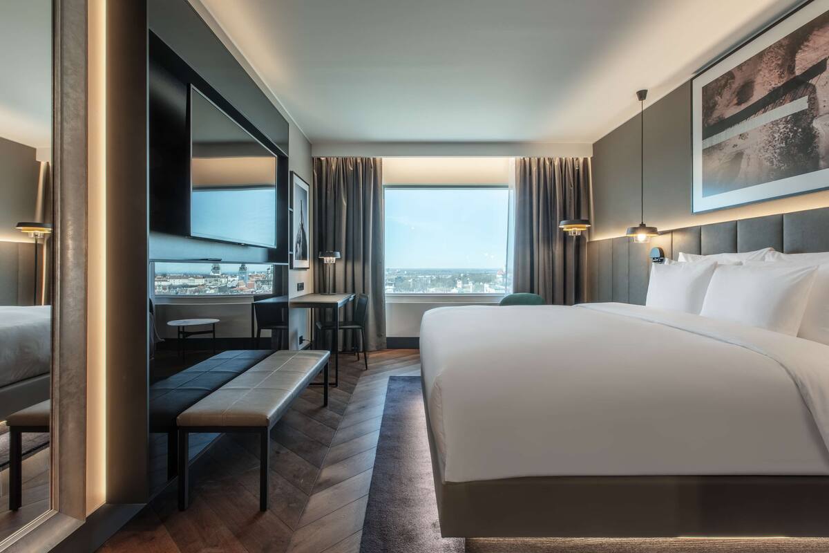 Quarto de hotel moderno com uma cama grande, uma escrivaninha, um banco e vista para a cidade.