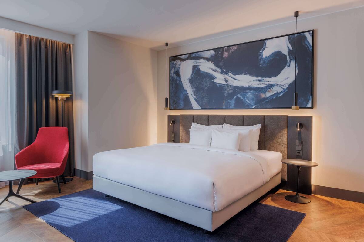 Quarto de hotel moderno com uma cama grande, uma cadeira vermelha, uma mesa pequena e um quadro abstrato grande na parede.