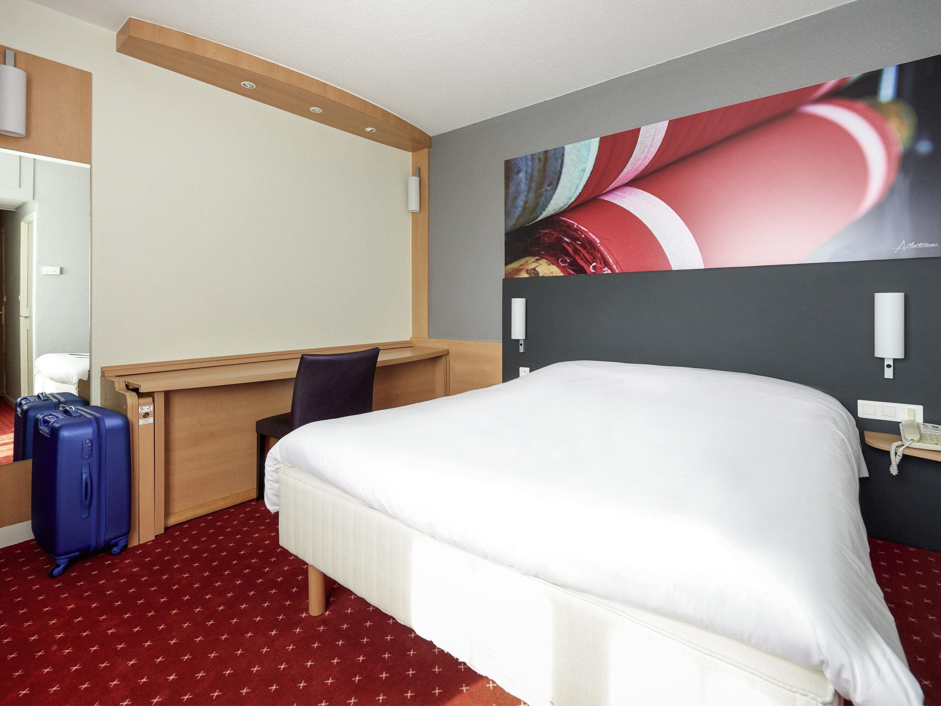 Quarto de hotel com uma cama, uma escrivaninha, uma cadeira, telefone e luminária na parede.