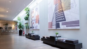 Um hall moderno com piso de madeira, balcão de recepção preto, plantas em vasos e grande obra de arte com vista urbana nas paredes.