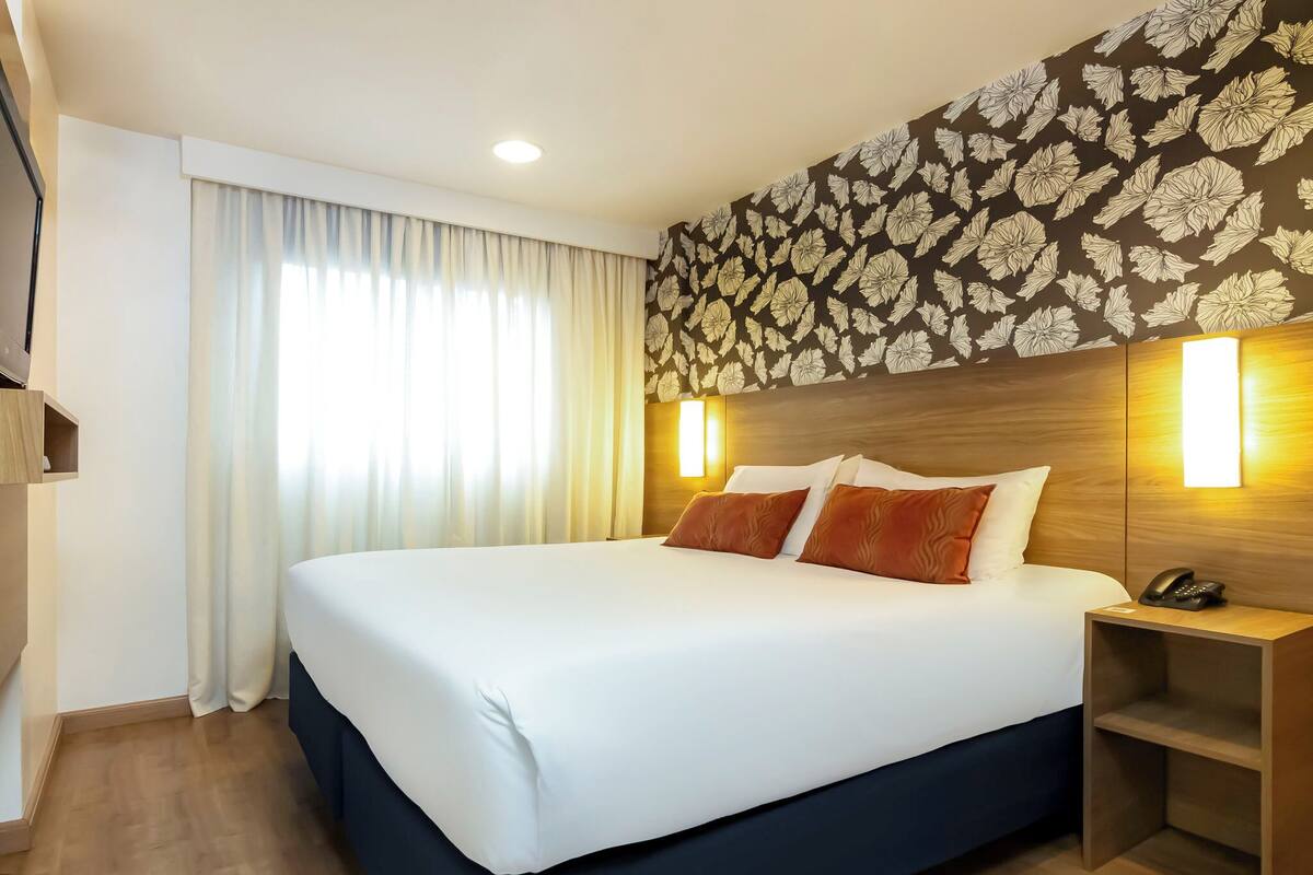 Quarto de hotel com cama grande, cabeceira de madeira, duas mesas de cabeceira e telefone fixado na parede.
