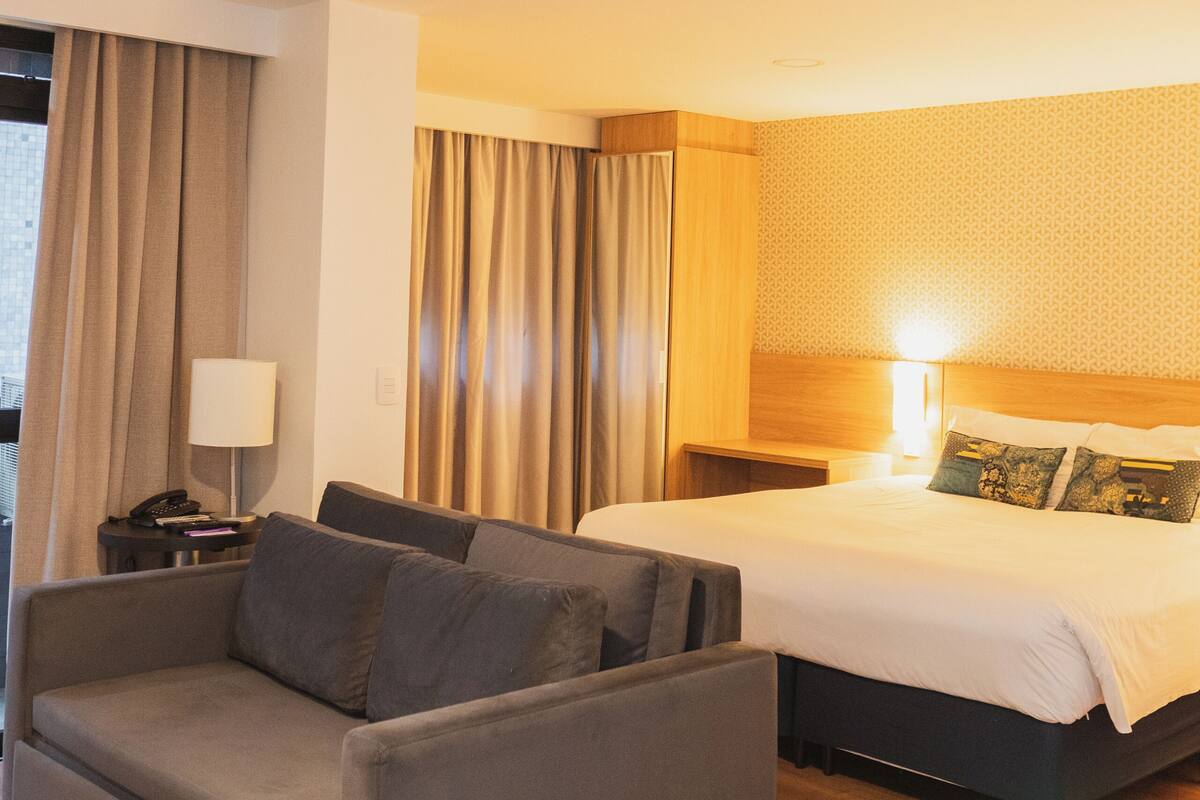 Quarto de hotel com cama, sofá, mesa de cabeceira com luminária e janela com cortinas.