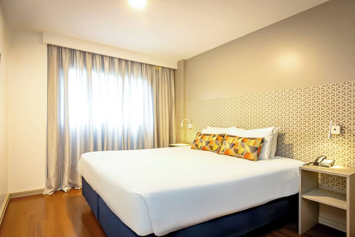 Quarto de hotel com cama grande, mesa de cabeceira, telefone fixado na parede e janela com cortinas.