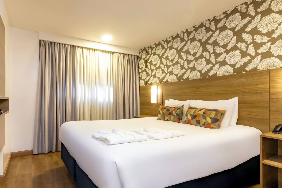 Quarto de hotel com cama bem arrumada, cabeceira de madeira, mesa de cabeceira com telefone e parede estampada.
