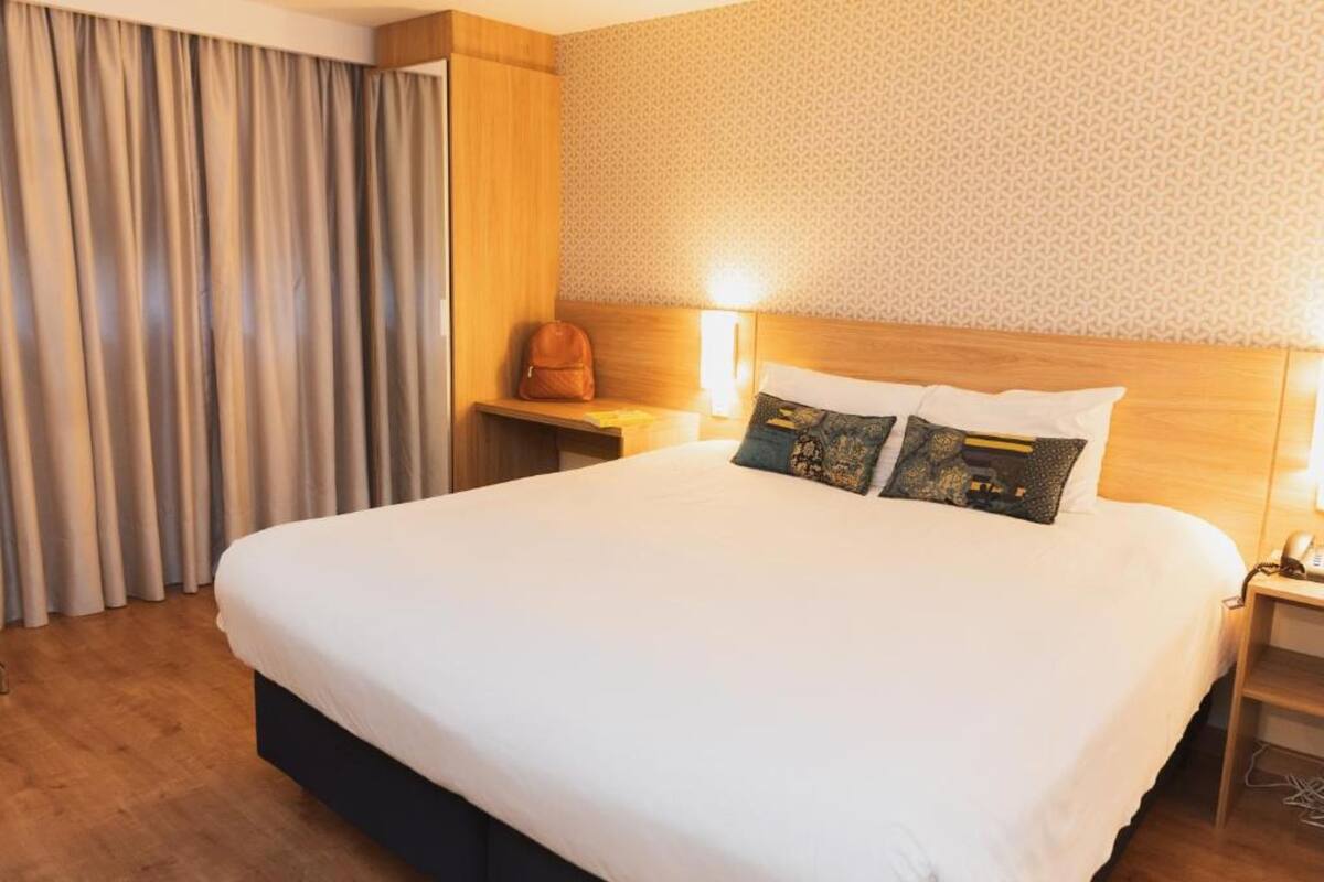 Quarto de hotel com uma cama grande, cabeceira de madeira, mesa de cabeceira com abajur, mala e parede estampada.