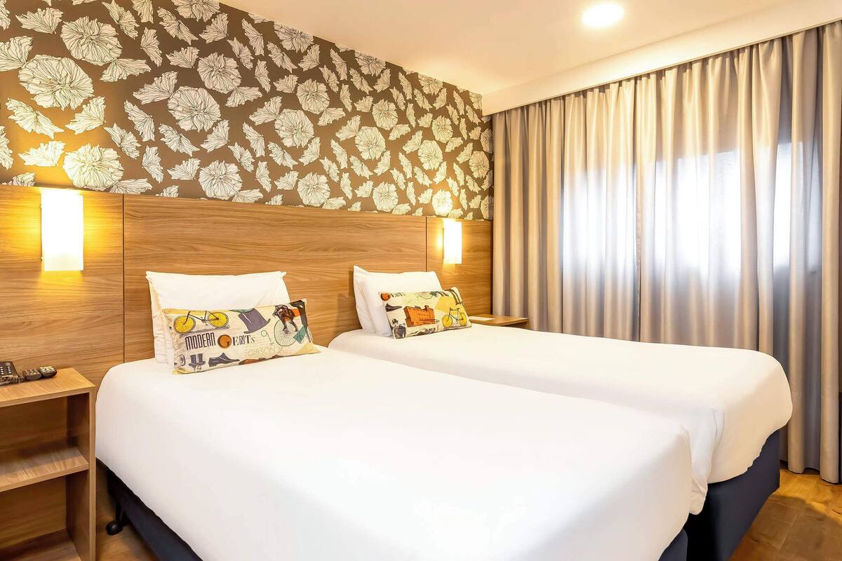 Quarto de hotel com duas camas, cabeceiras de madeira, papel de parede floral e uma janela grande com cortinas.