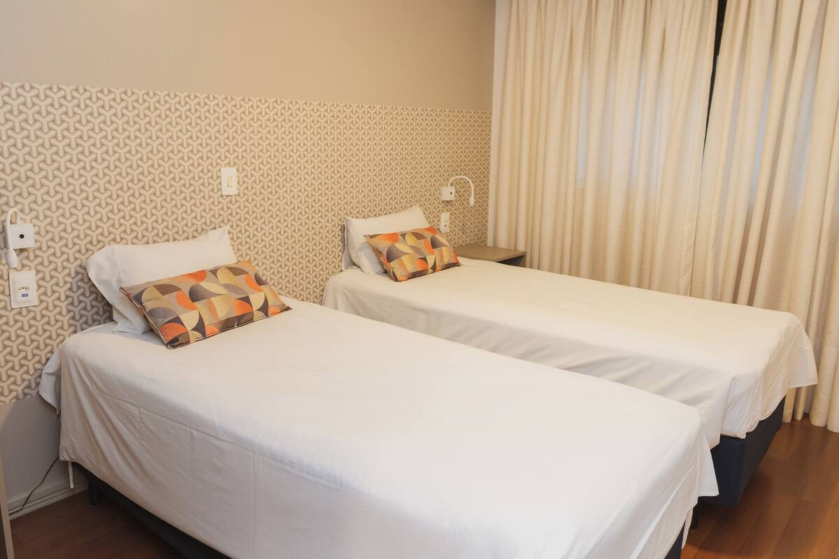 Quarto de hotel com duas camas de solteiro, cada uma com colcha branca e almofadas estampadas. O quarto possui papel de parede estampado e uma janela com cortinas.