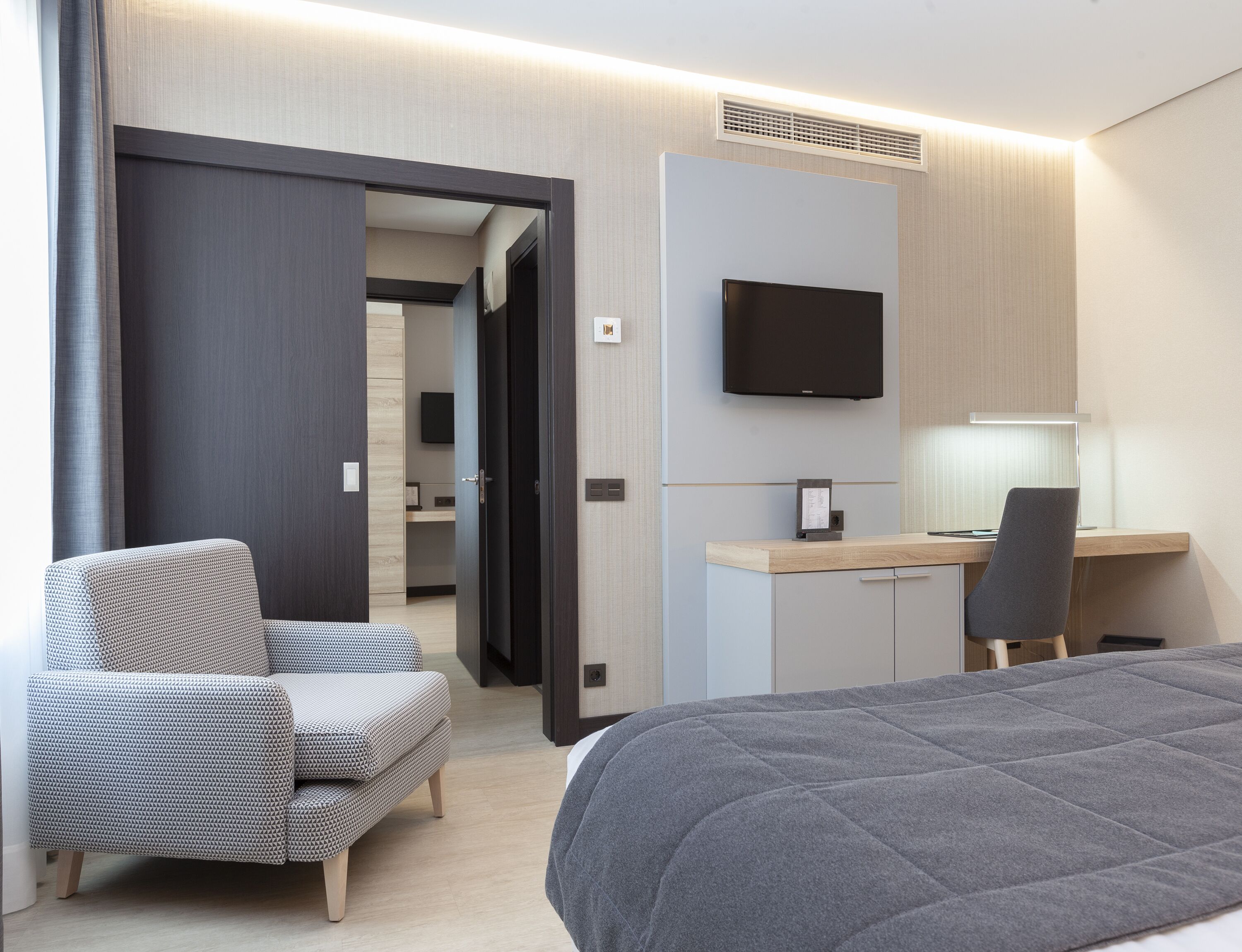 Quarto de hotel com cama, escrivaninha, cadeira, televisão e poltrona estampada.