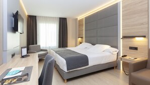 Quarto de hotel moderno com uma cama grande, uma escrivaninha com cadeira, uma televisão de tela plana na parede e uma mesa de cabeceira com telefone e abajur.